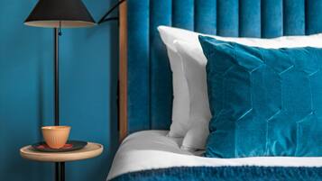 Quarto com parede azul, cabeceira de madeira, cama com colcha azul e branca, abajur de chão preto e mesa redonda pequena com uma tigela.