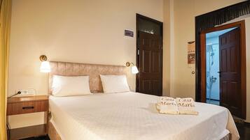 Cama bem arrumada com lençóis brancos e duas toalhas com o nome "Casa Maria", mesa de cabeceira de madeira com uma luminária, e uma porta que dá para o banheiro.