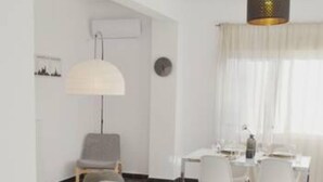 Uma sala de estar moderna com um sofá cinzento, uma mesa redonda pequena e uma área de refeições ao fundo.