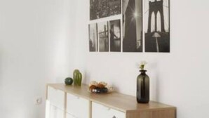 Uma sala de estar moderna com um sofá cinzento, uma mesa consola de madeira com objetos decorativos e uma parede com uma série de fotografias a preto e branco.