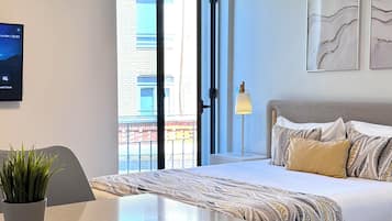 Quarto de hotel com cama, escrivaninha, planta em vaso, abajur e janela com vista para edifícios.