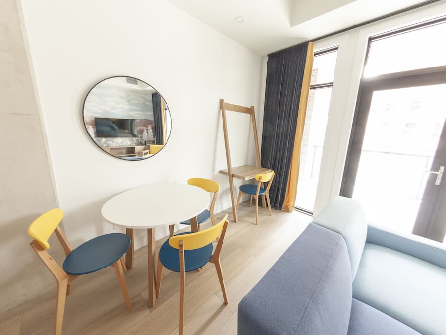 Quarto moderno com um espelho redondo, uma mesa redonda e duas cadeiras com almofadas amarelas e azuis. Há um sofá com uma almofada azul e uma janela com cortinas.
