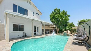 Uma piscina com uma casa branca ao fundo, uma área de varanda coberta e uma cadeira suspensa à beira da piscina.
