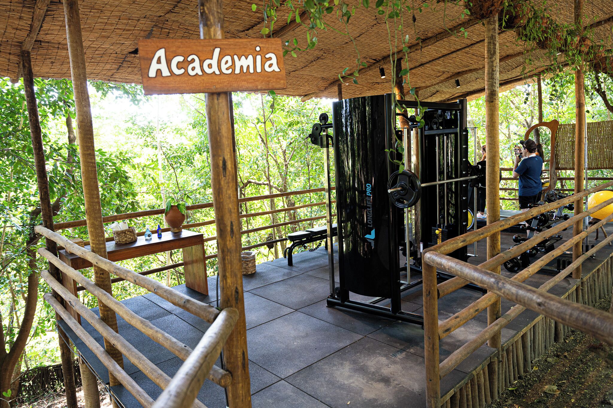 Área de ginástica ao ar livre com equipamentos de exercício, uma placa de madeira com a inscrição 'Academia' e um cenário natural.