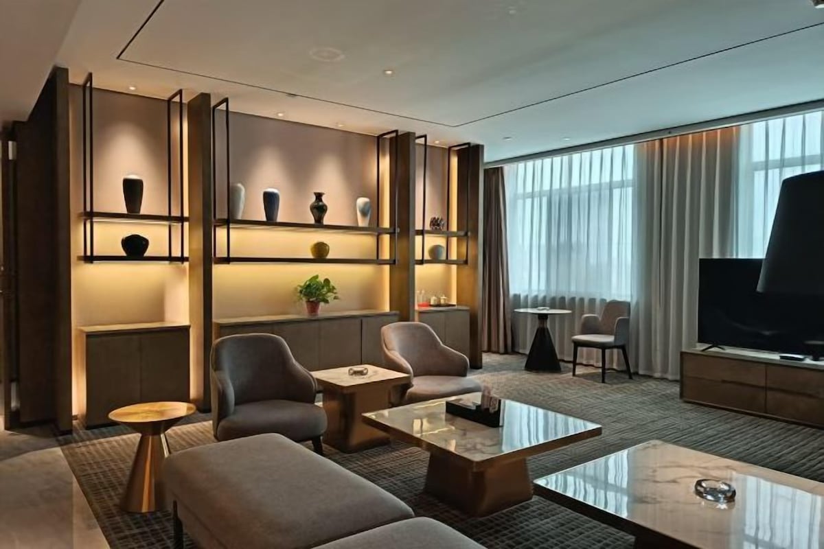 Quarto de hotel moderno com sofá, poltronas, mesa de centro, televisão e estante de livros.