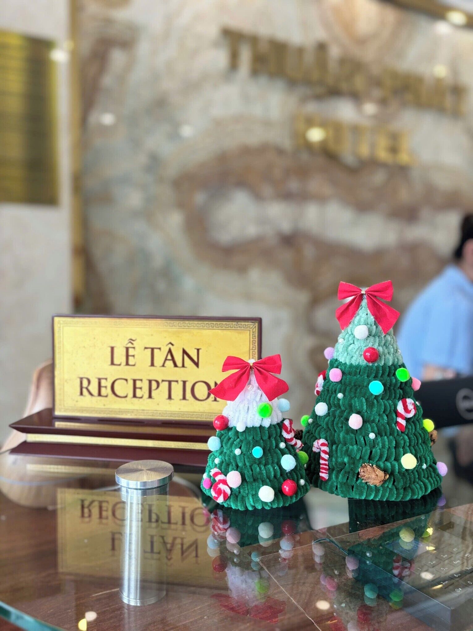 Um balcão de recepção com uma placa indicando "Lễ Tấn Reception" e duas pequenas árvores de Natal com enfeites coloridos.