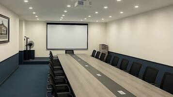 Sala de conferências com uma mesa comprida, cadeiras, tela de projetor e um aparelho de ar condicionado.
