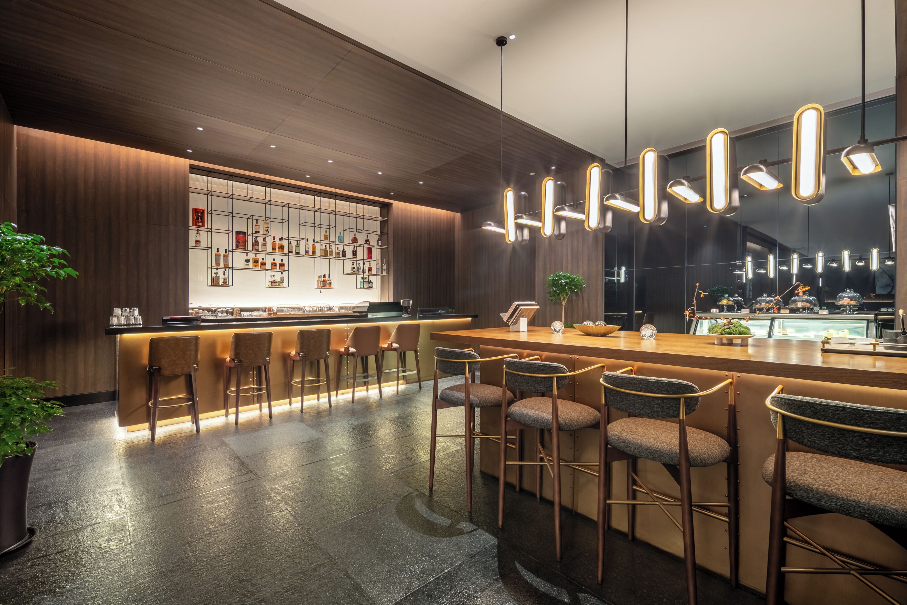 Uma área de bar moderna com balcão de madeira, banquetas, iluminação pendente e uma parede espelhada refletindo o bar.