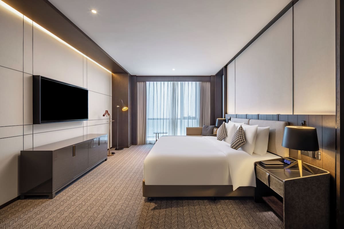 Quarto de hotel moderno com uma cama grande, televisão de tela plana, mesa de cabeceira com abajur e vista para a cidade através de grandes janelas.