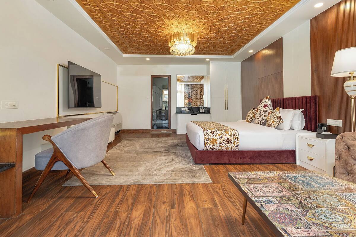 Quarto de hotel com teto de madeira, cama com colcha estampada, escrivaninha com cadeira, televisão e abajur.