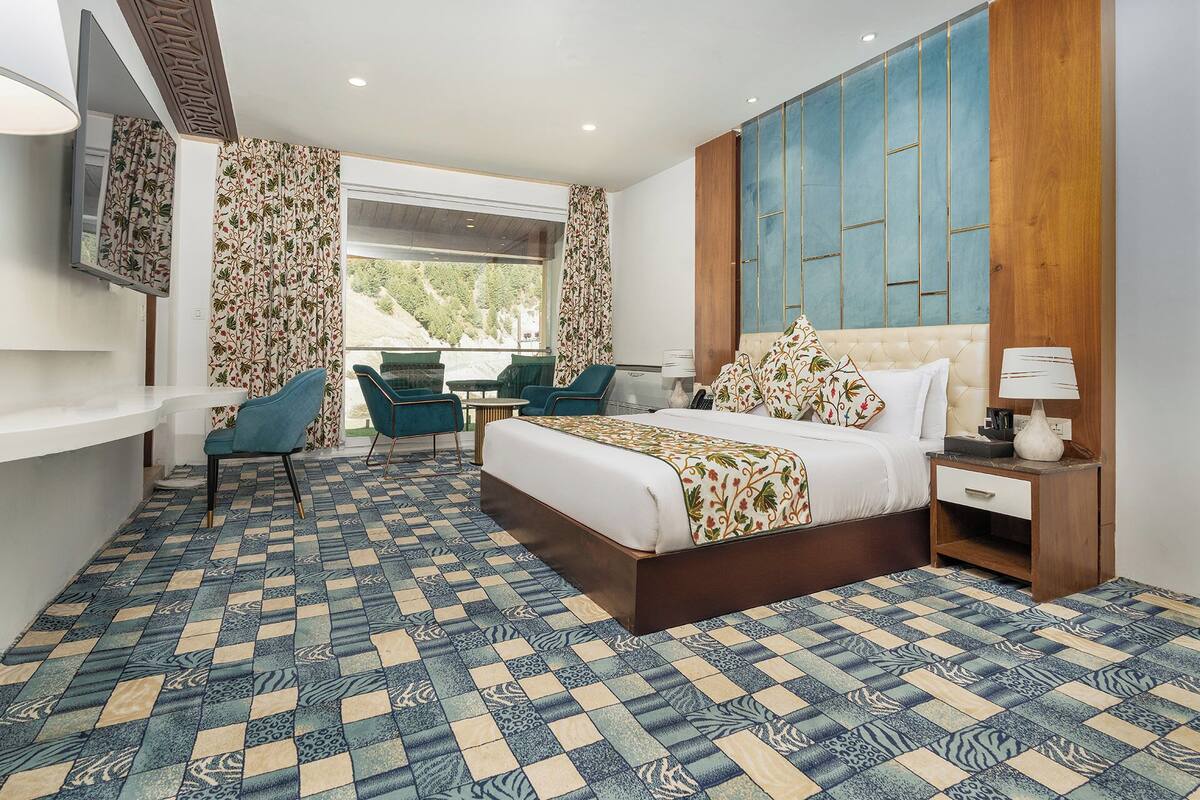 Quarto de hotel com uma cama, duas cadeiras, uma escrivaninha, mesa de cabeceira e uma janela grande.
