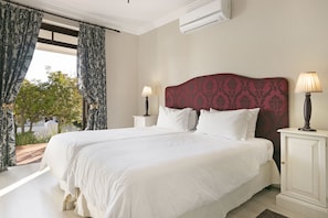 Uma cama grande com roupa de cama branca, cabeceira vermelha, abajures e vista para o verde lá fora.