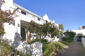 Um edifício branco com varanda, caminho pavimentado e vegetação exuberante.