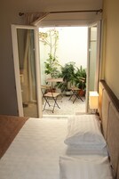Quarto de hotel com cama, uma pequena mesa, cadeiras e vista para uma varanda com plantas em vasos.