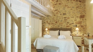 Quarto com parede de pedra, cama com roupa branca, duas mesas de cabeceira com abajures e um pequeno banco.