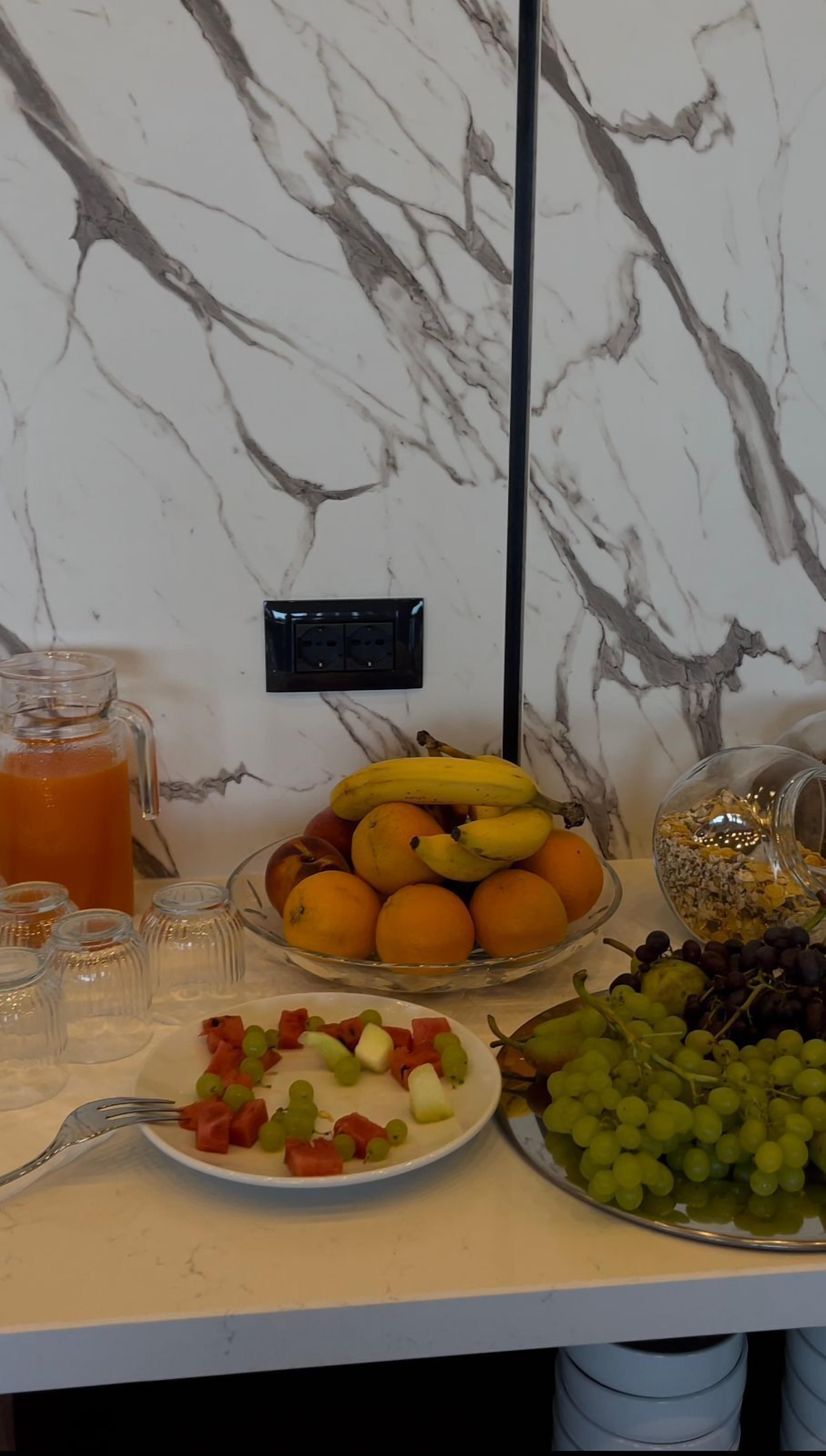 Um buffet de café da manhã com frutas, um jarro de suco de laranja e uma tomada na parede.