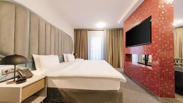 Quarto de hotel com uma cama, uma escrivaninha com telefone e luminária, uma TV fixada numa parede com azulejos vermelhos e uma janela com cortinas.