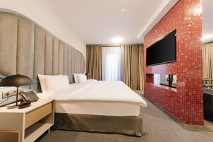 Quarto de hotel com uma cama, uma escrivaninha com telefone e luminária, uma TV fixada numa parede com azulejos vermelhos e uma janela com cortinas.