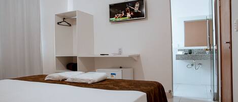 Quarto de hotel com cama, televisão presa à parede, guarda-roupa e banheiro com pia e espelho.
