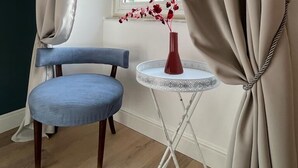 Um quarto com uma cadeira azul, uma mesa redonda pequena com um vaso de flores vermelhas e cortinas bege.