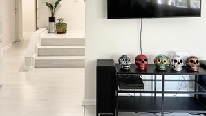 Uma sala de estar com uma TV de tela plana na parede, um móvel preto com decoração de caveiras, uma escada que leva a outro cômodo e um vaso com planta.