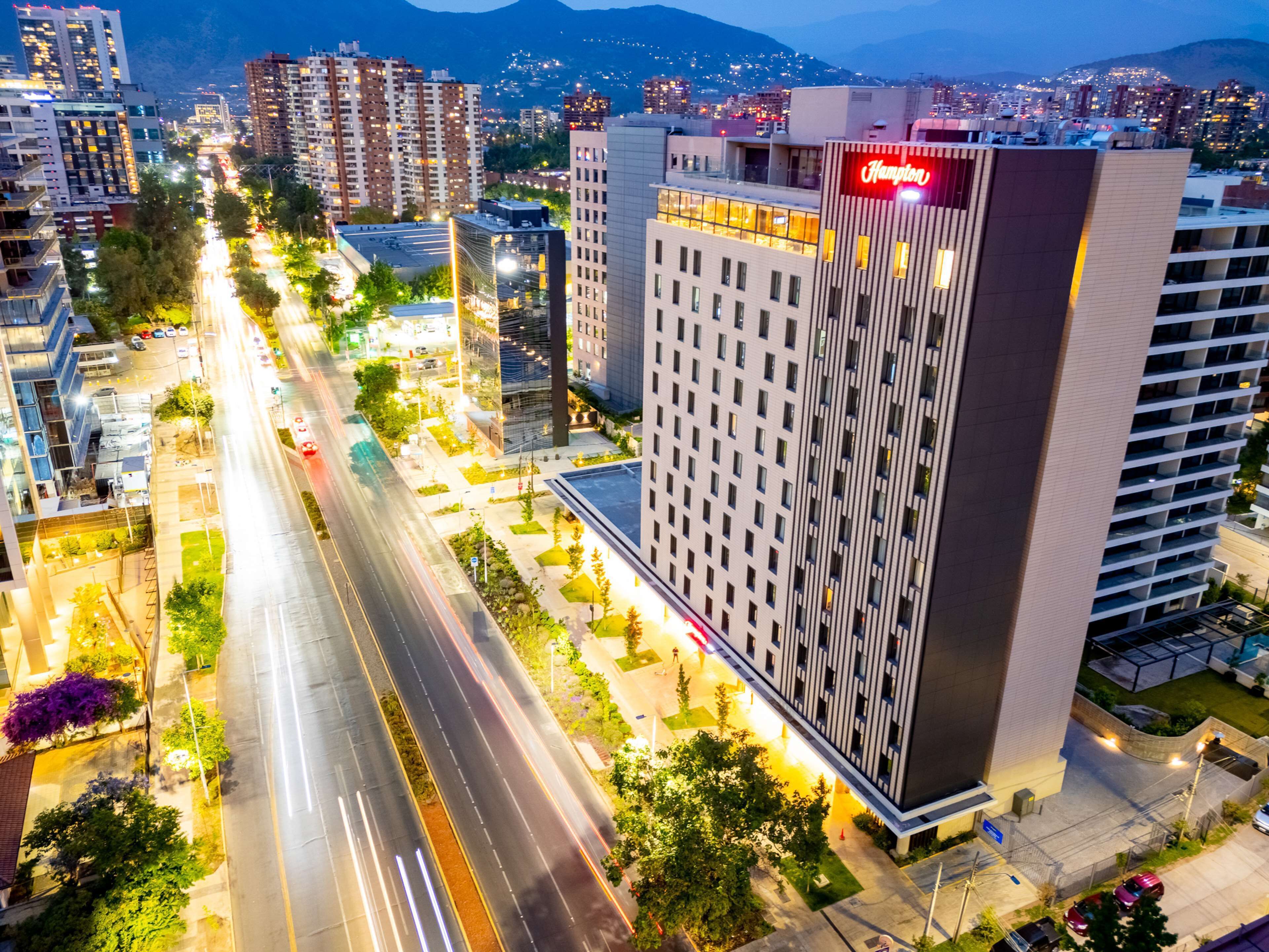 Um hotel moderno com um grande letreiro 'Hampton', situado numa área urbana bem iluminada com ruas e áreas verdes visíveis.