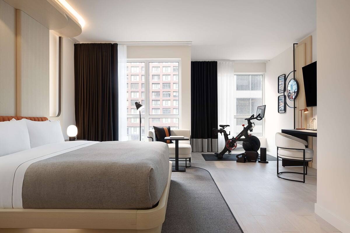 Quarto de hotel com uma cama, uma escrivaninha, uma bicicleta ergométrica e vista para edifícios pelas janelas.