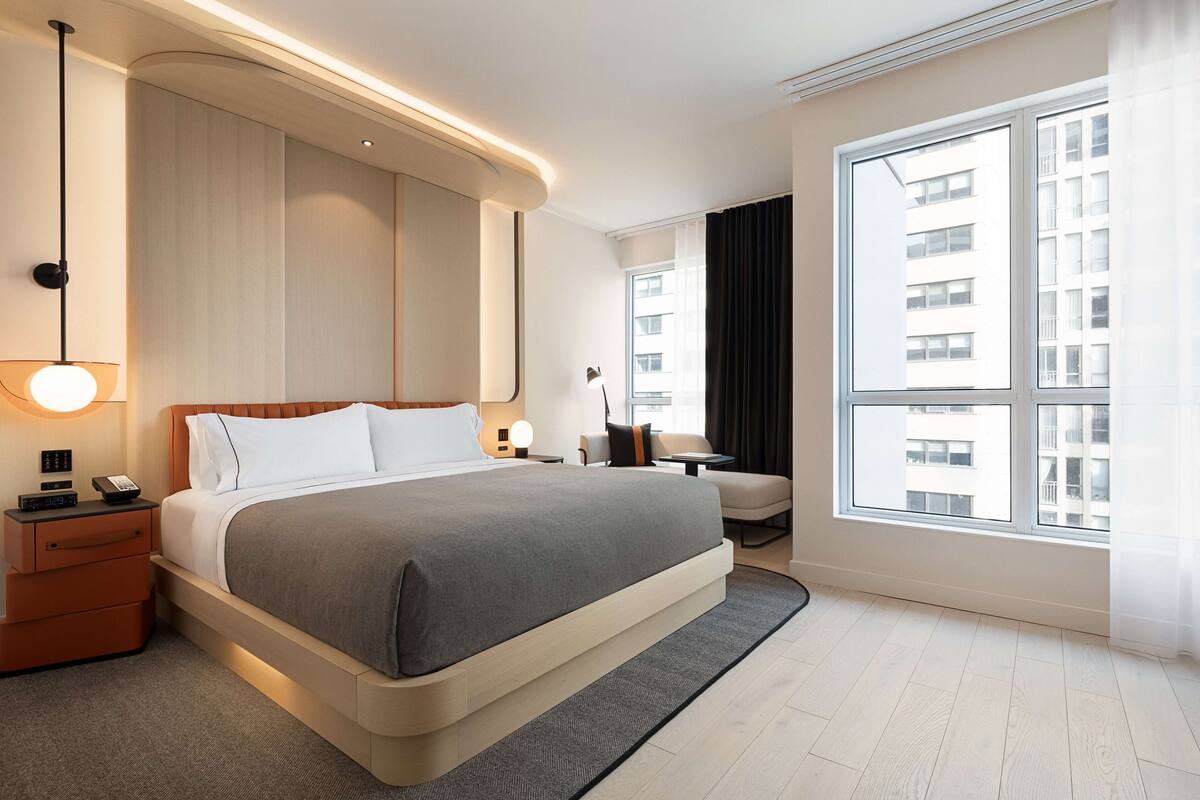 Quarto de hotel moderno com cama grande, mesas de cabeceira, escrivaninha e uma janela ampla com vista para a cidade.