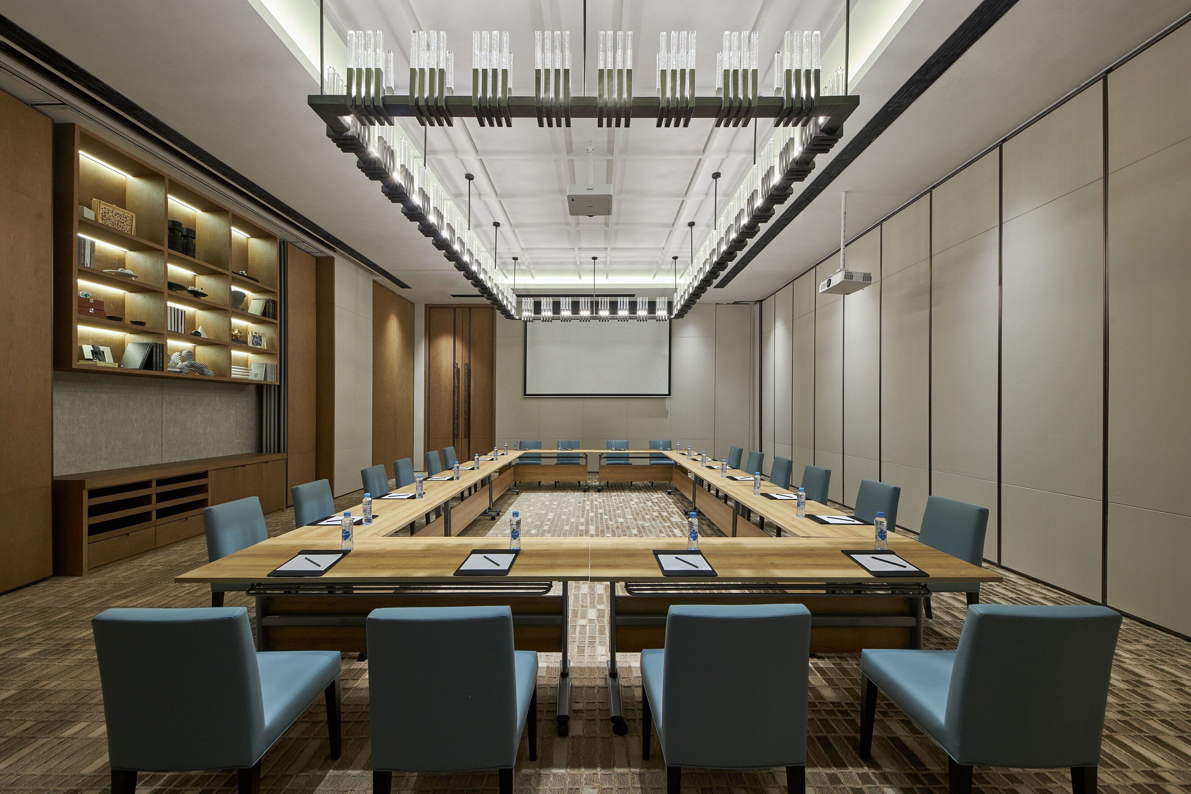 Uma sala de conferência com uma mesa comprida, cadeiras azuis e uma tela de projeção.