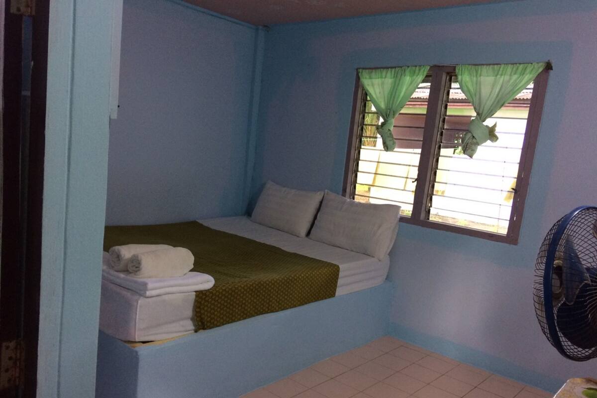 Quarto pequeno com cama de solteiro, janela com cortina verde, colcha branca e toalha branca sobre a cama.
