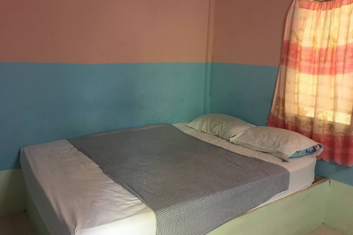 Uma cama de solteiro com um cobertor cinza e branco xadrez, um travesseiro branco e uma cortina colorida.