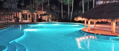 Uma piscina noturna com borda curva, rodeada por palmeiras e uma estrutura com telhado de palha.