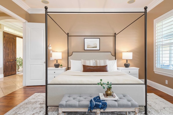 Uma cama com dossel sobre quatro postes, com roupa de cama branca e um travesseiro marrom, com mesas de cabeceira com luminárias de cada lado, em um quarto com piso de madeira e uma grande janela com persianas.