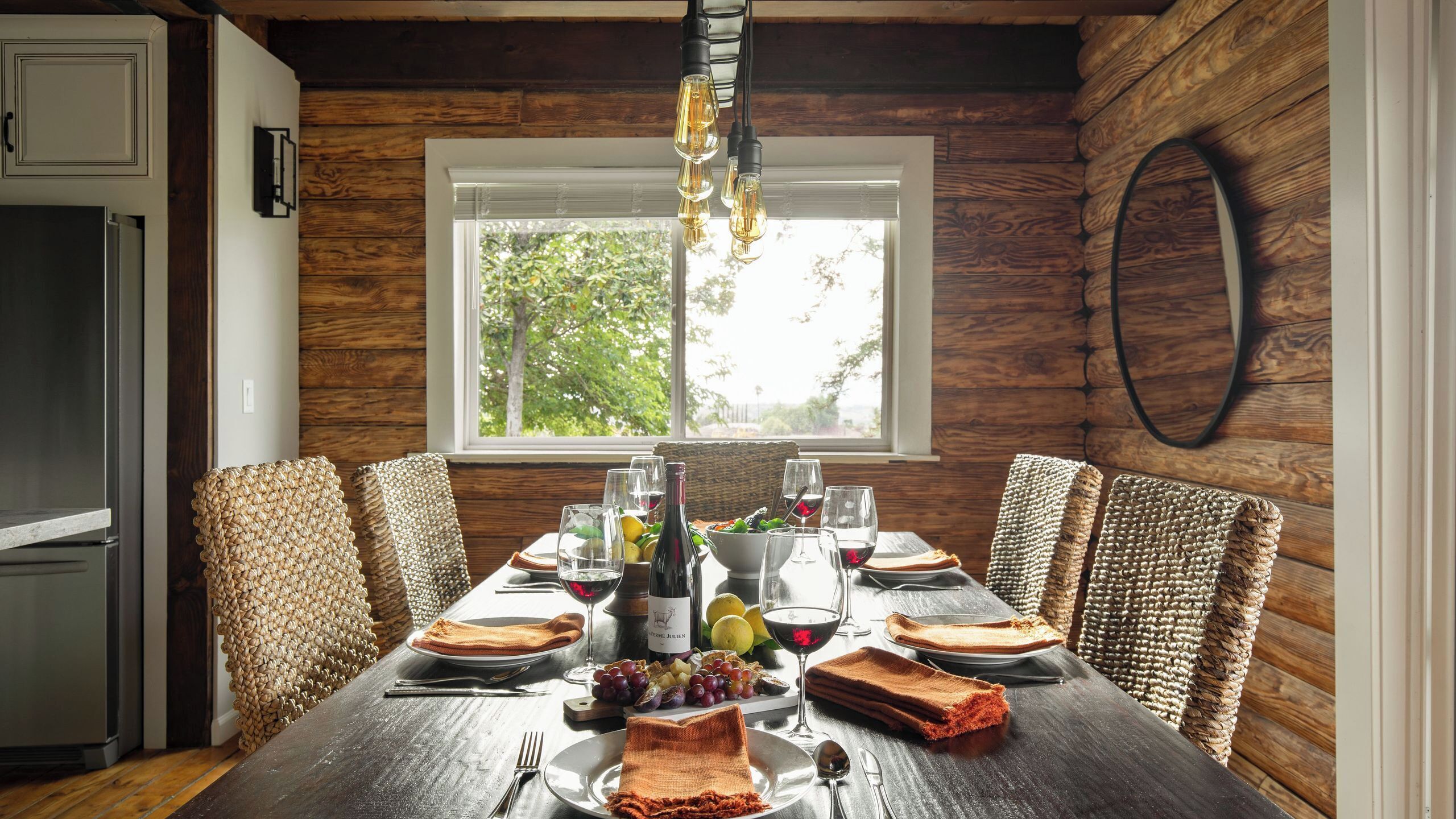Uma sala de jantar com uma mesa de madeira posta para seis pessoas, rodeada por cadeiras de fibra, vista para a janela e um lustre.