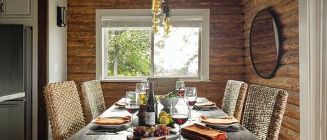 Uma sala de jantar com uma mesa de madeira posta para seis pessoas, rodeada por cadeiras de fibra, vista para a janela e um lustre.