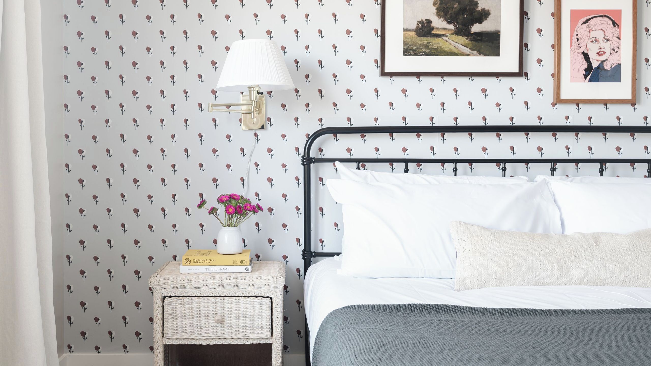 Quarto com cama, mesa de cabeceira com abajur, quadro emoldurado e parede com padrão floral.