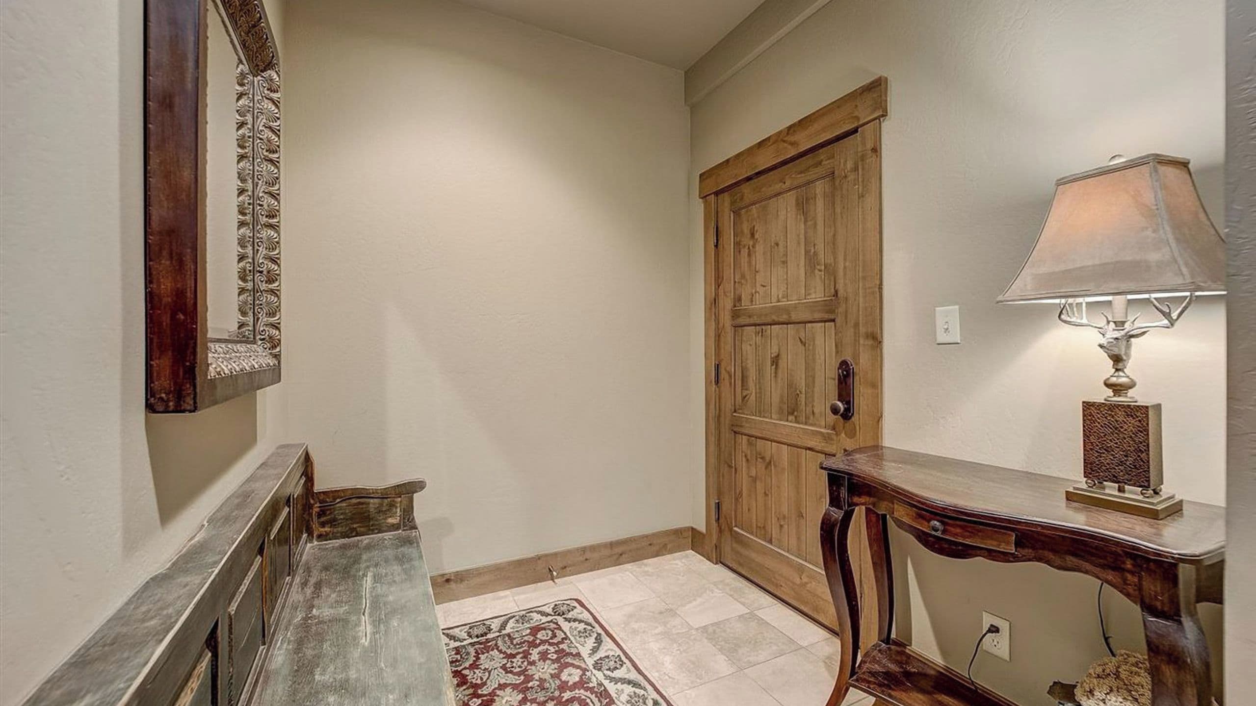 Um corredor com porta de madeira, um espelho, uma luminária e um banco.