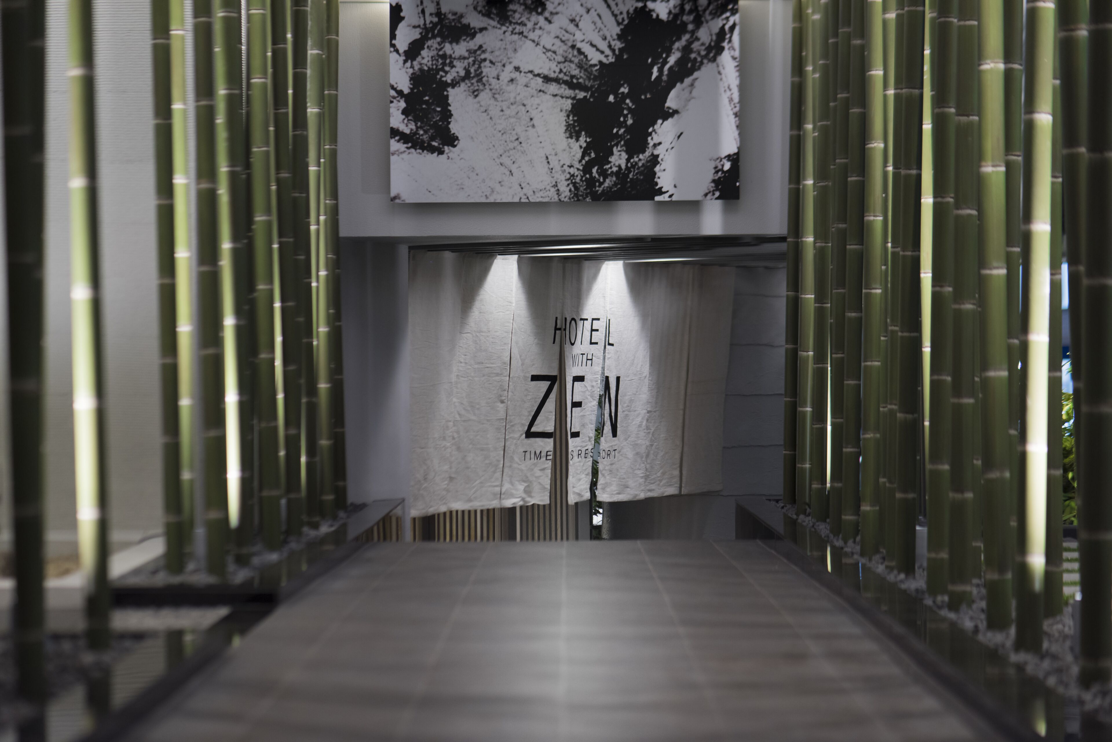 Um corredor com estruturas verticais semelhantes a bambu, uma placa na parede com a inscrição "HOTEL WITH ZEN TIM-ES RESORT" e um piso com padrão claro.