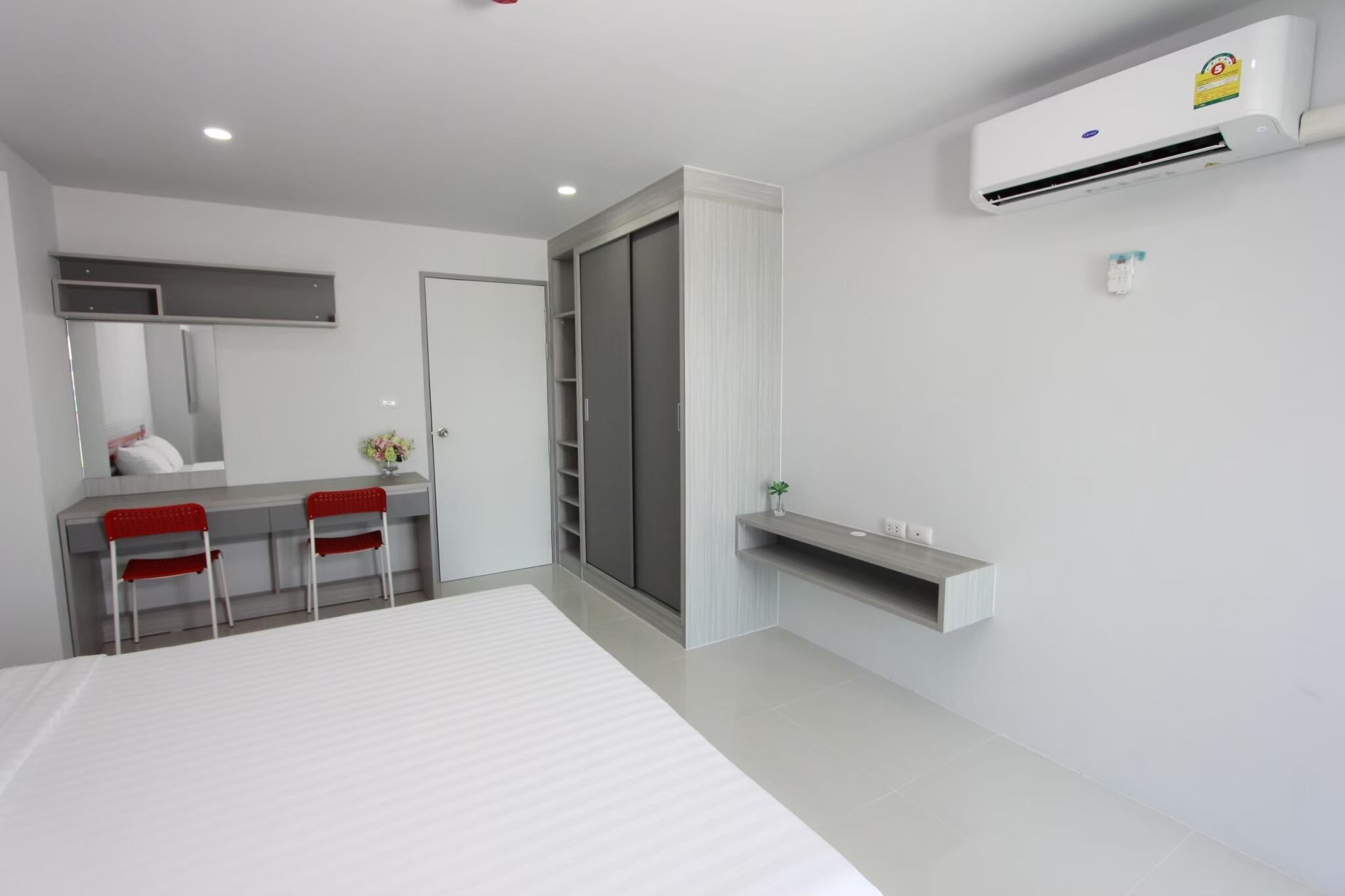 Quarto de hotel moderno com uma cama, uma mesa com duas cadeiras vermelhas, um guarda-roupa e um aparelho de ar condicionado.