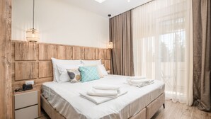 Quarto com cabeceira de madeira, cama com roupa de cama branca, mesa de cabeceira com abajur e janela com cortinas translúcidas.
