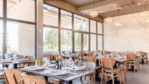 Restaurante com várias mesas e cadeiras, janelas amplas e iluminação pendente.