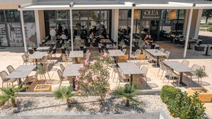 Uma área externa com mesas e cadeiras, um pequeno jardim com plantas e o restaurante OLEA Restaurant ao fundo.