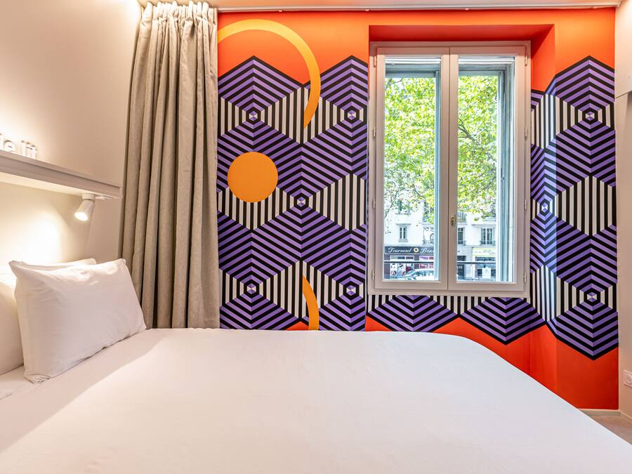 Quarto de hotel com cama, cortinas, janela e vista, e parede com padrão geométrico.