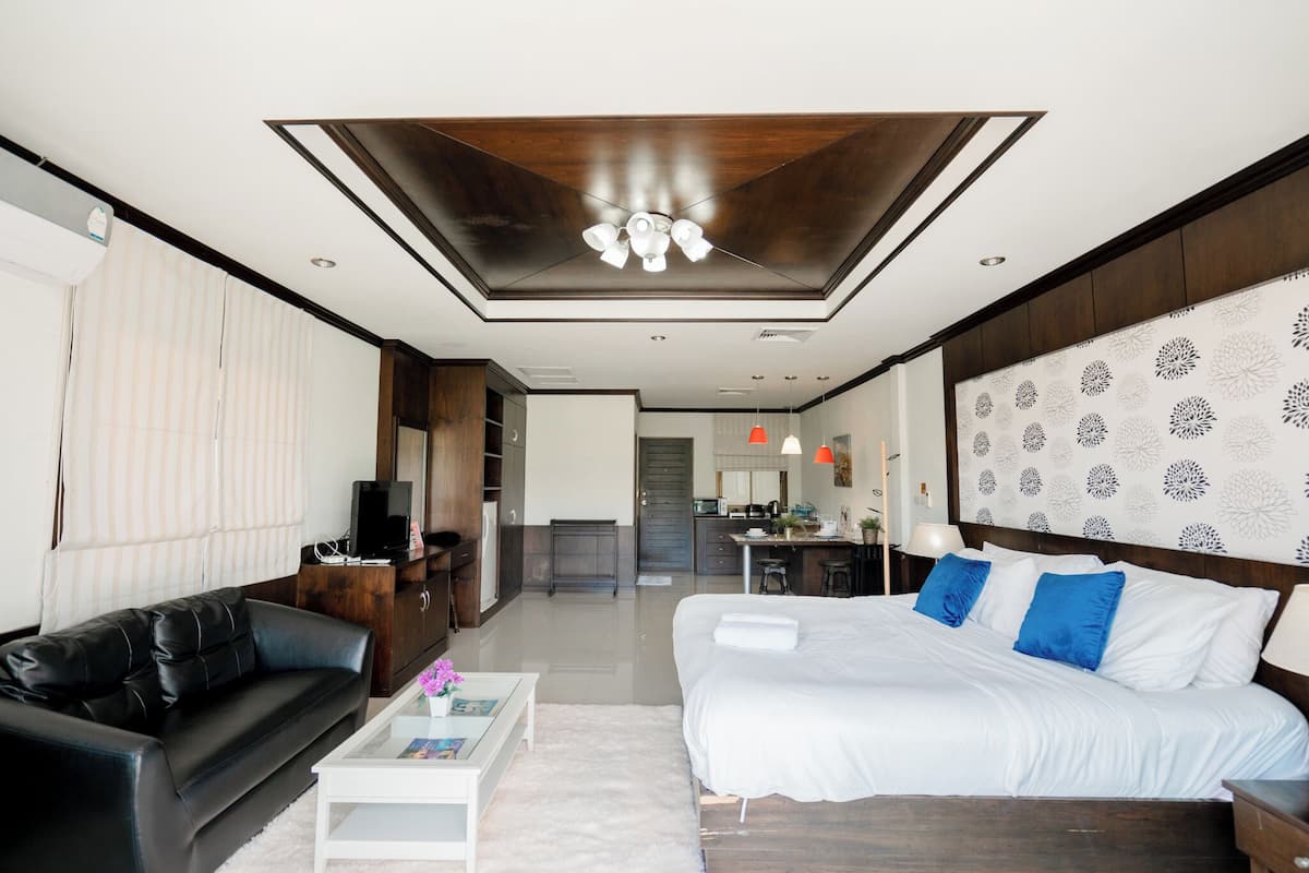Quarto de hotel moderno com uma cama grande, um sofá de couro preto, uma pequena mesa de centro e uma área de cozinha ao fundo.
