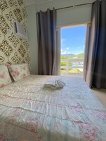 Quarto com cama, roupa de cama estampada com flores, janela com cortinas e vista para uma piscina e montanhas.