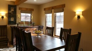 Uma área de refeições com uma mesa de madeira, cadeiras e uma tigela de frutas. Há duas janelas com cortinas, teto de madeira e luminárias fixadas na parede.