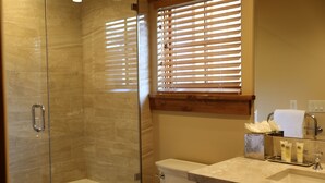 Casa de banho com cabine de chuveiro de vidro, sanita, lavatório com bancada de mármore e janela com estores.