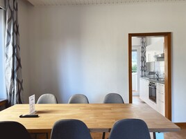 Uma sala de jantar moderna com uma mesa de madeira e cadeiras cinzas estofadas. Uma placa é visível sobre a mesa, e cozinha é visível através de uma porta aberta.