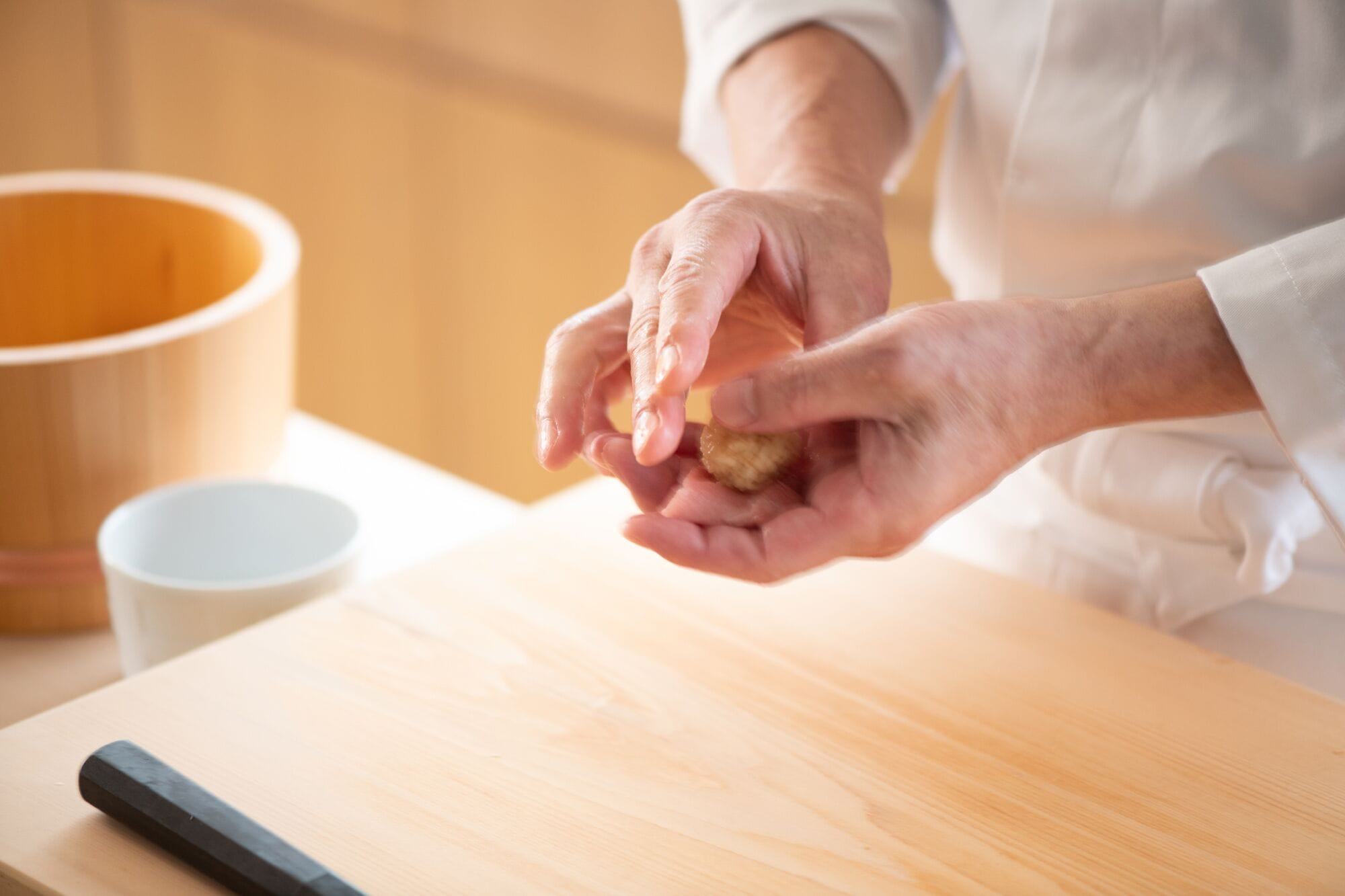 Uma pessoa segurando um objeto pequeno, possivelmente um biscoito, sobre uma mesa de madeira com uma xícara branca e um objeto preto por perto.