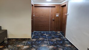 Um corredor com porta de madeira identificada com o número 402, chão de azulejos com padrão de peixe e um relógio digital marcando 21:12.